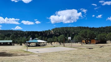 Campo aberto com um pavilhão, quadra de basquete e uma colina arborizada ao fundo.