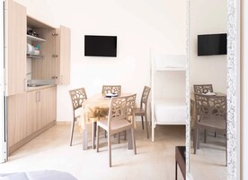 Quarto compacto com cozinha americana, mesa de jantar com cadeiras, televisão de tela plana na parede e uma cama branca.