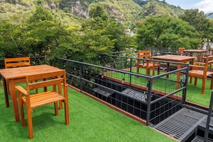 Uma área de estar ao ar livre com mesas e cadeiras de madeira, cercada por um guarda-corpo de metal, com vista para grama verde e árvores.