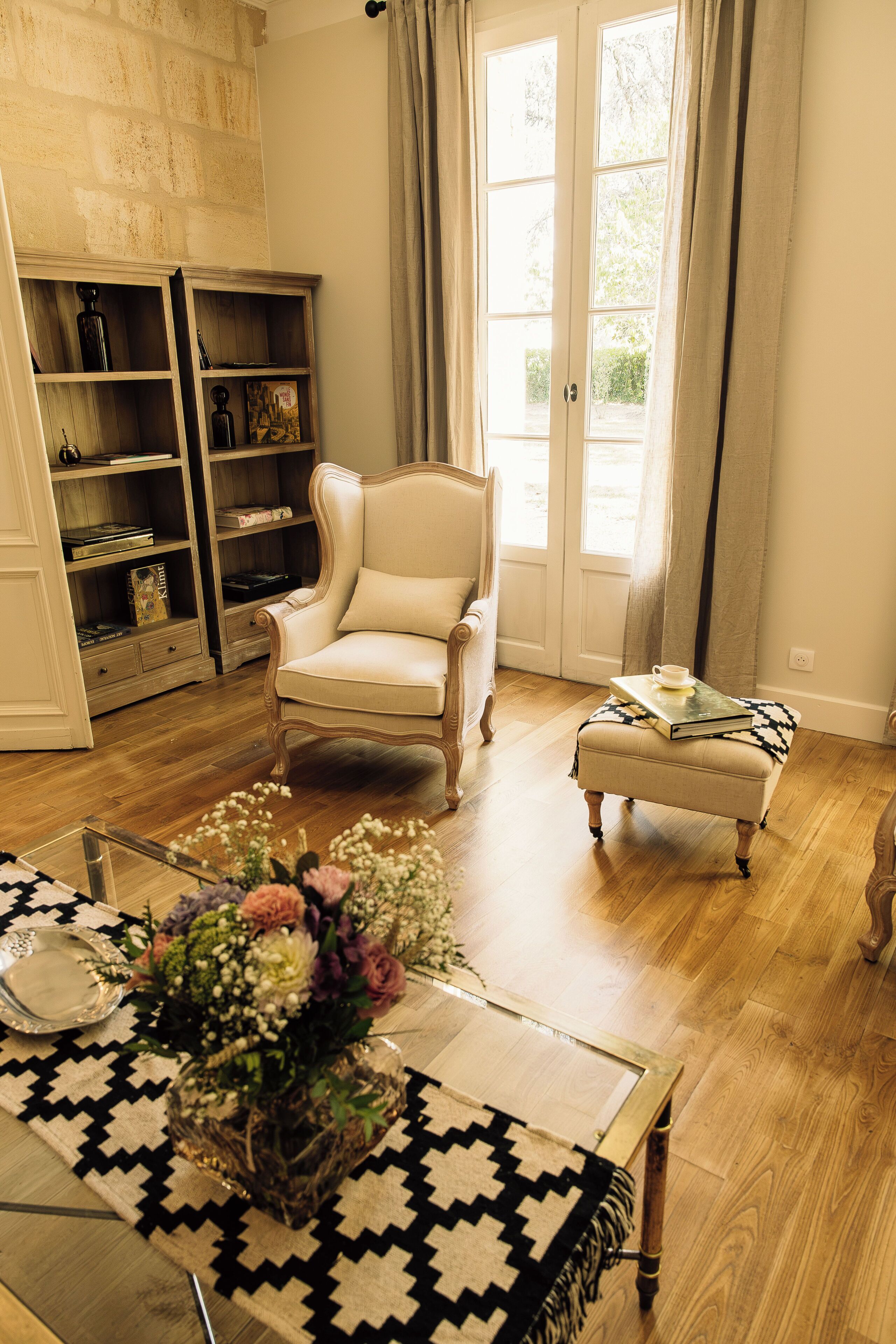 Um quarto com piso de madeira, uma poltrona branca, uma mesinha com um vaso de flores e uma estante de livros.