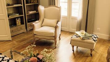 Um quarto com piso de madeira, uma poltrona branca, uma mesinha com um vaso de flores e uma estante de livros.
