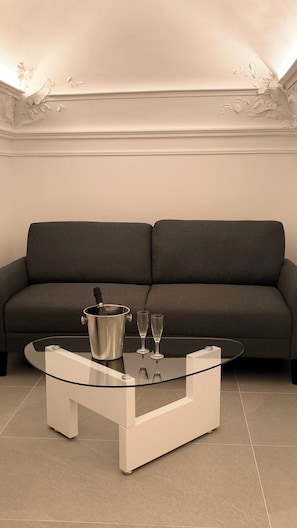Uma sala de estar moderna com um sofá cinza escuro, uma mesa de centro de vidro com um recipiente de metal e copos, e um piso de azulejos.