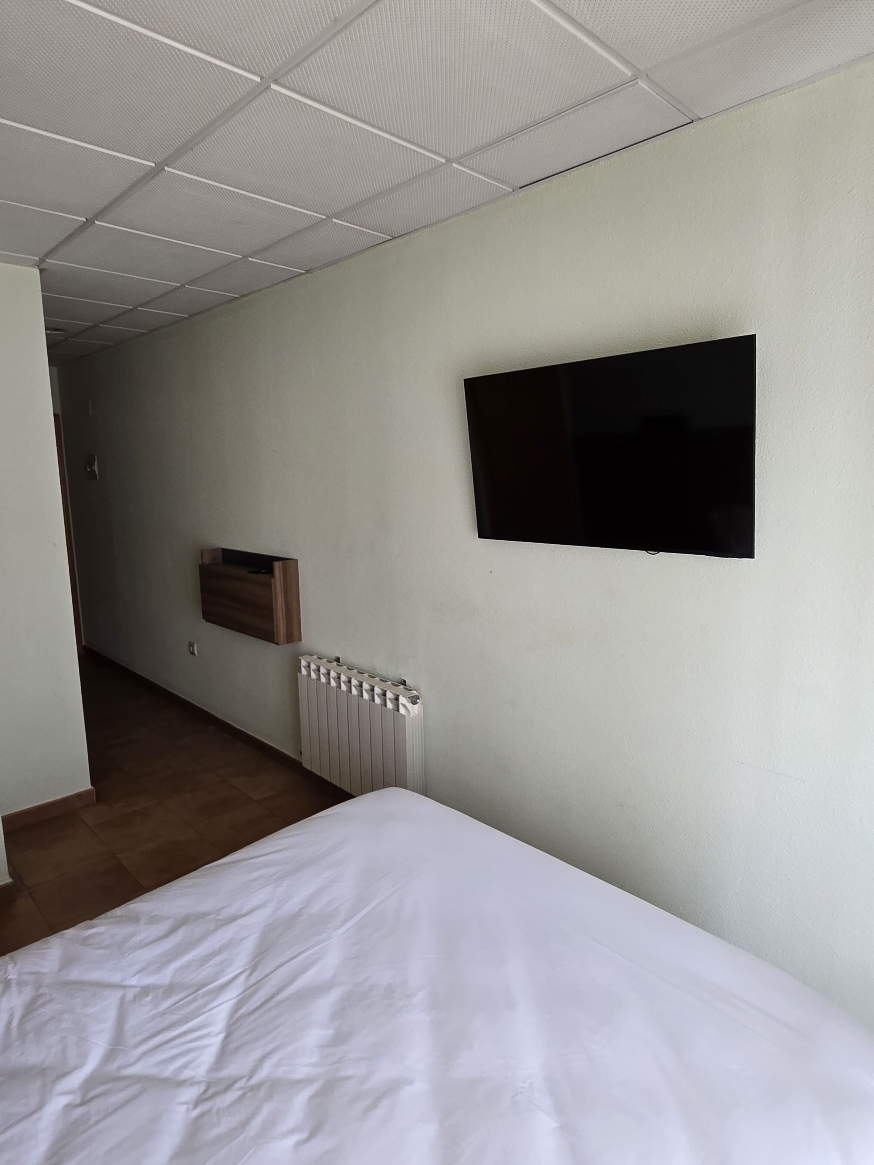 Quarto de hotel com cama, televisão na parede, radiador e armário de madeira.