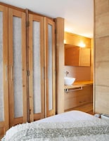 Quarto com cama, guarda-roupa de madeira com portas espelhadas, mesa de cabeceira e banheiro com pia e espelho.