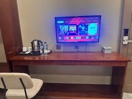 Uma mesa de madeira com uma televisão de tela plana exibindo a interface de um serviço de streaming, uma chaleira, duas garrafas de água e uma cadeira.