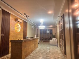 Uma área de recepção de hotel com piso de mármore, balcão de atendimento, um lustre e uma janela grande.