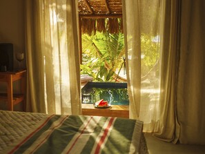 Quarto com cama, cortinas, uma mesinha com um copo e vista para o verde lá fora.