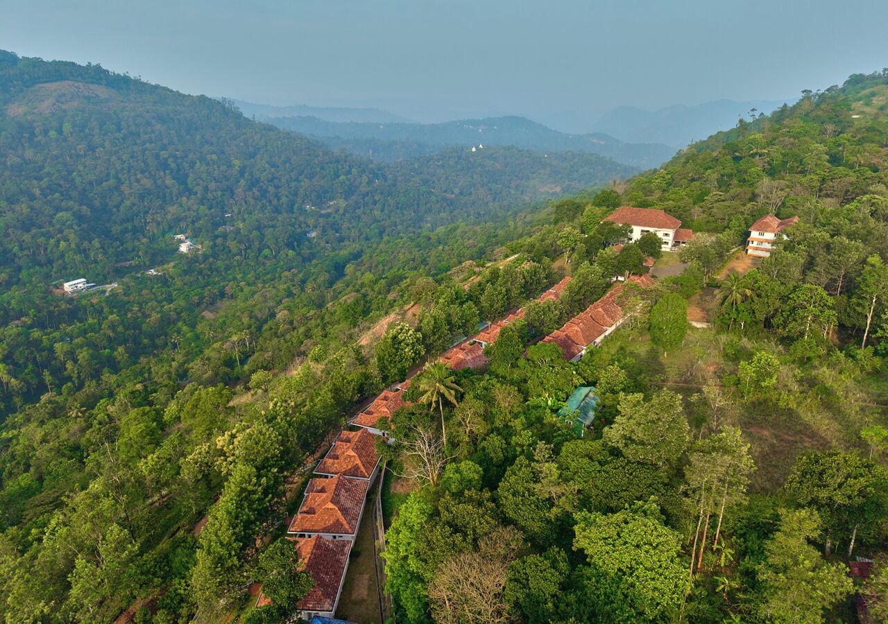 Um edifício comprido e estreito com telhado vermelho, rodeado por uma floresta verde e colinas.