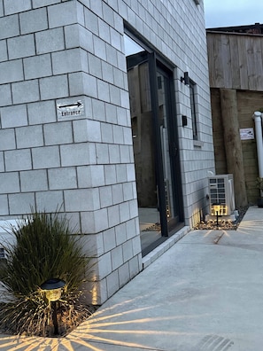 Entrada de um edifício com placa indicando a direção, uma porta e um pequeno acessório de iluminação externa.