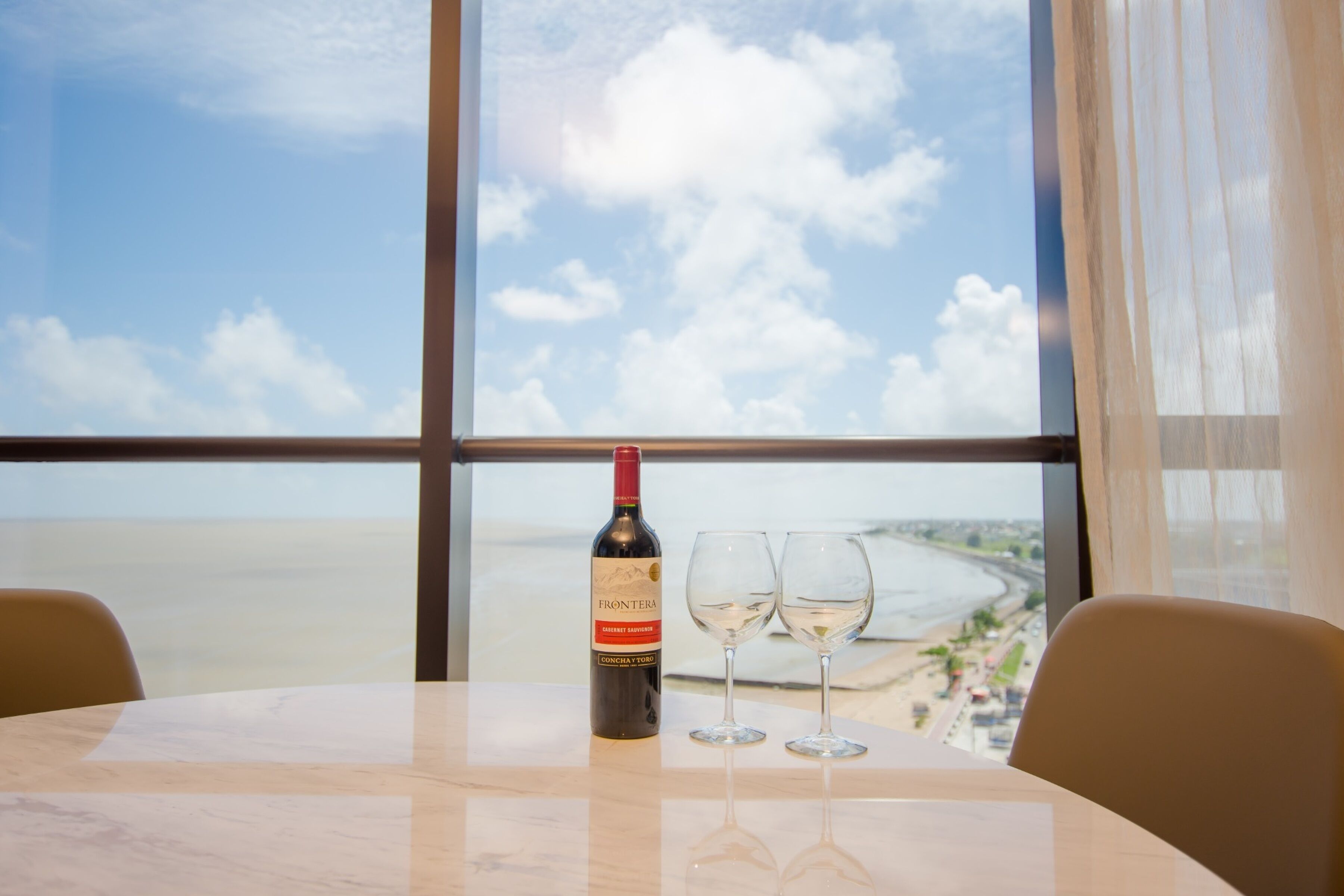 Uma mesa com uma garrafa de vinho e duas taças, vista para o mar através de grandes janelas.
