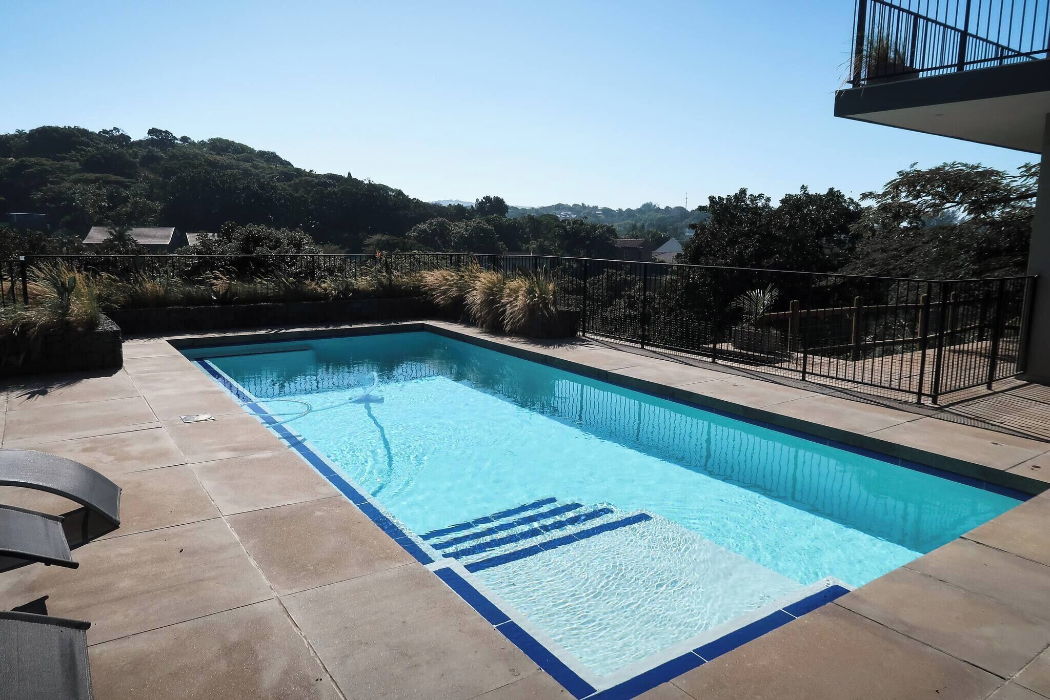 Uma piscina retangular com água azul cristalina, rodeada por um deck revestido de azulejos e uma cerca de metal.