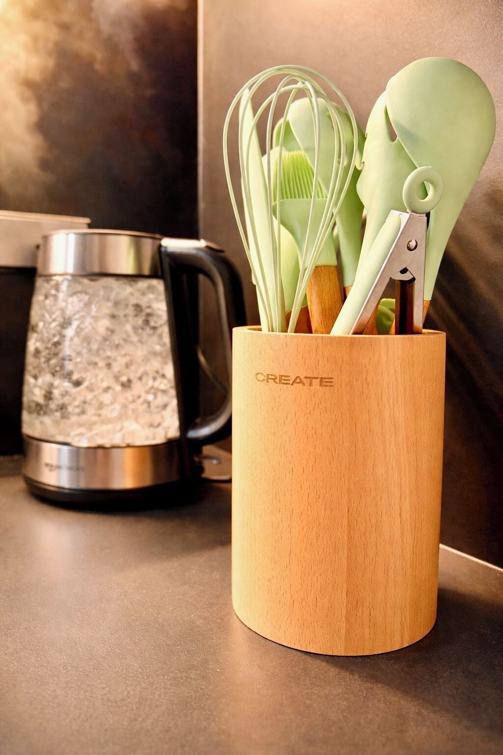 Uma caneca de madeira com utensílios de cozinha, uma chaleira de aço inoxidável e um recipiente texturizado sobre uma bancada.