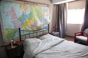 Um quarto com um mapa grande na parede, uma cama, uma cadeira e uma escrivaninha com uma luminária.