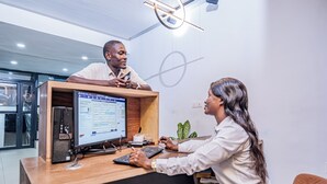 Bancada de recepção com monitor de computador, teclado e uma xícara de café. Há uma planta na mesa e uma pessoa sentada em uma cadeira.