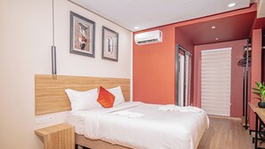 Quarto de hotel com cama de madeira, roupa de cama branca, pequena mesa de cabeceira, ar condicionado e um quadro na parede.