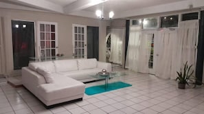 Um sofá branco, uma mesa de centro de vidro e uma planta em vaso numa sala de estar com pavimento em mosaico e porta de vidro.