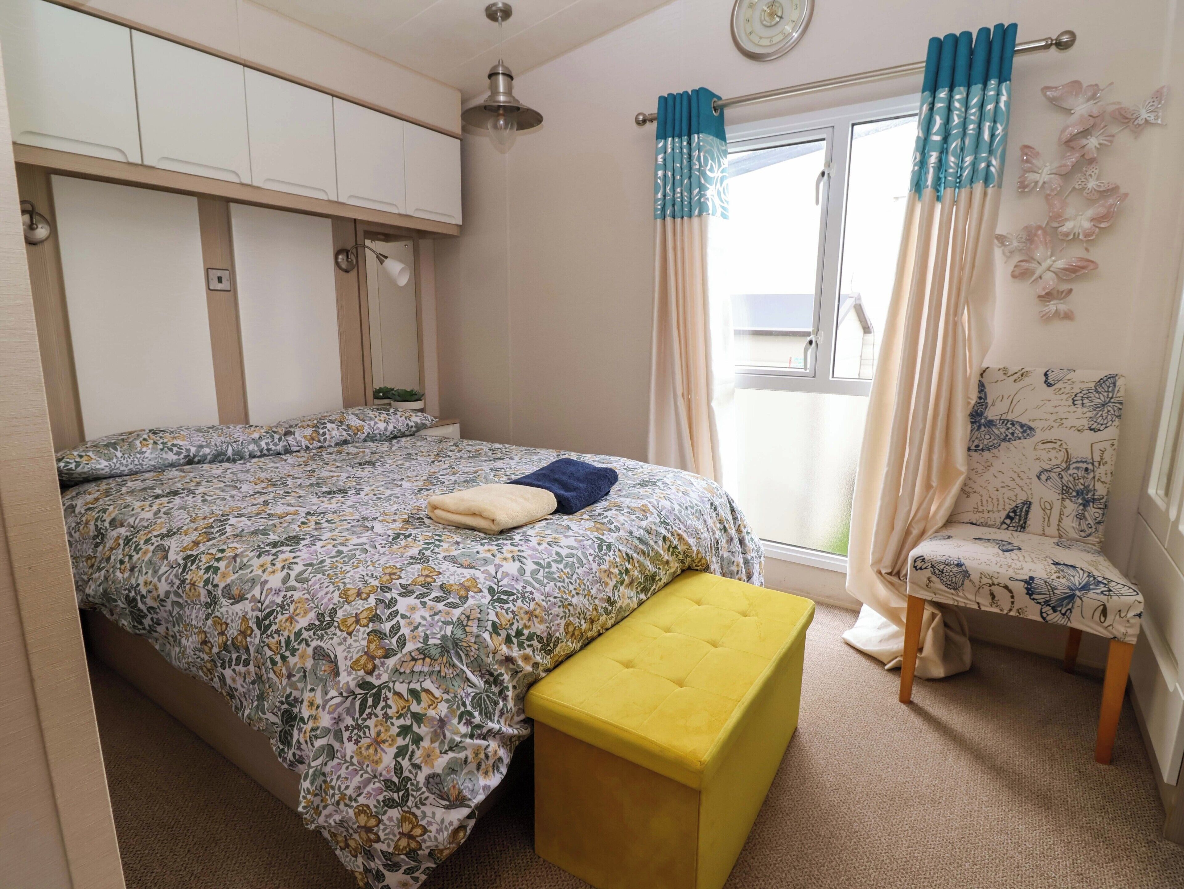 Quarto com cama, um banquinho, uma cadeira, janela com cortinas, relógio e ventilador de teto.