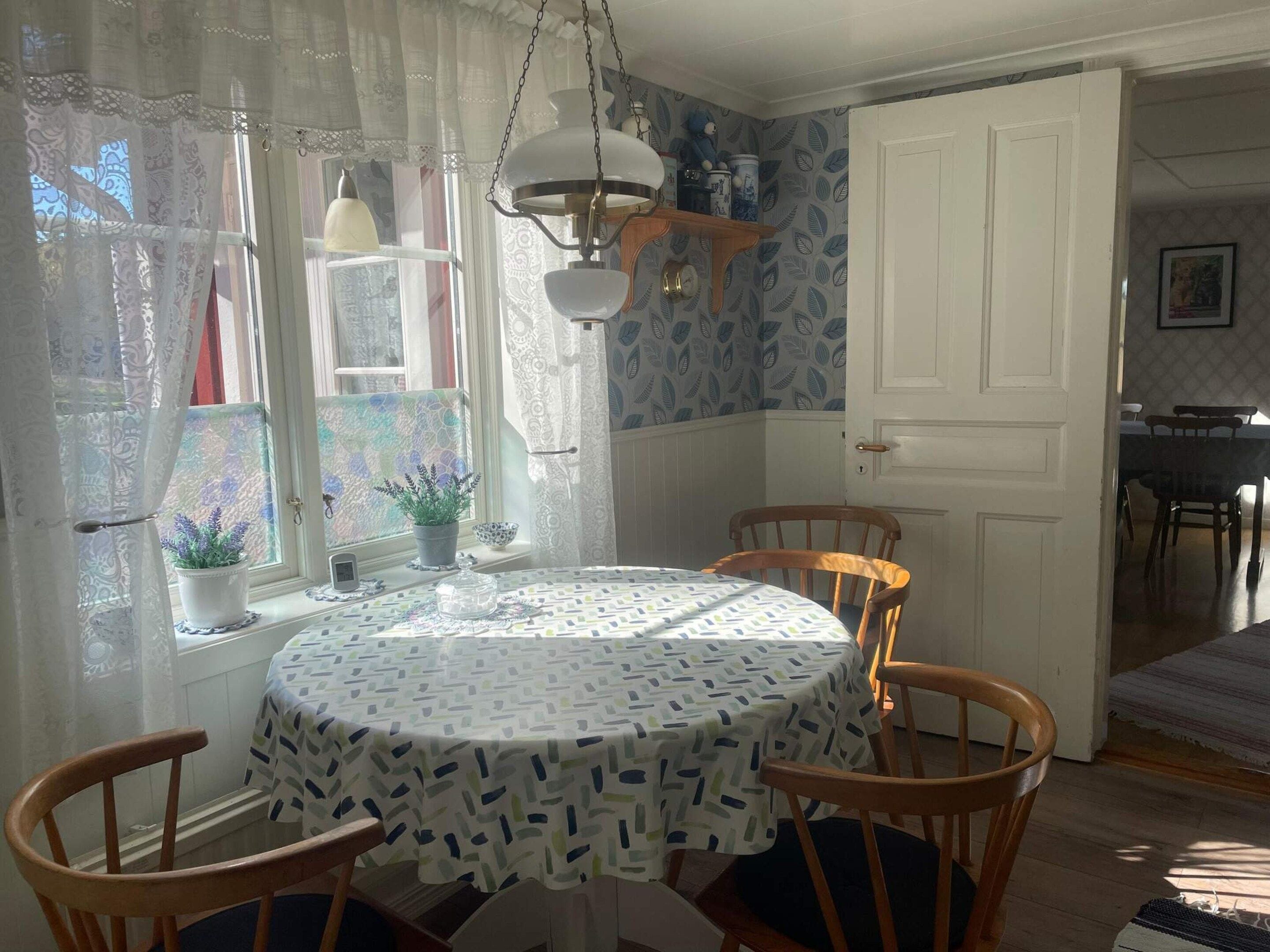 Uma sala de jantar com mesa redonda, cadeiras, janela com cortinas de renda e parede estampada.