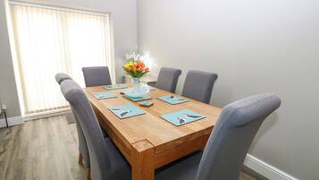 Uma sala de jantar com uma mesa de madeira, seis cadeiras estofadas cinzentas, um vaso com flores e uma janela com persianas.