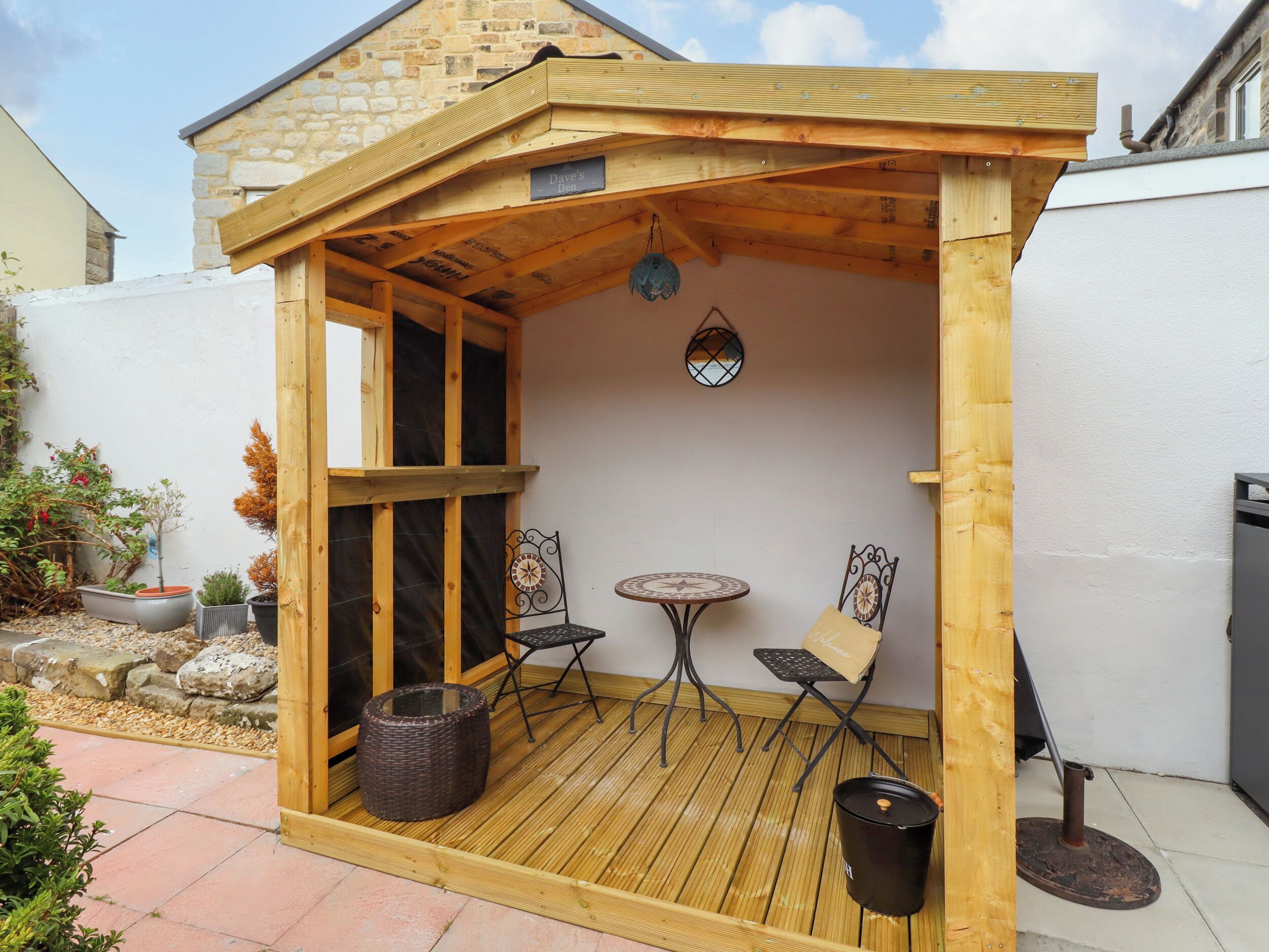Uma pérgola de madeira com uma mesa, cadeiras, uma luminária pendente e um pequeno vaso com planta.