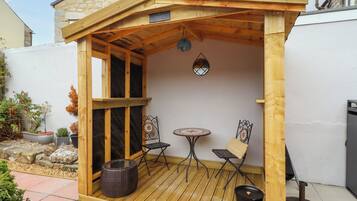 Uma pérgola de madeira com uma mesa, cadeiras, uma luminária pendente e um pequeno vaso com planta.