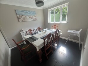 Uma sala de jantar com uma mesa posta para quatro pessoas, cadeiras de madeira, um quadro na parede e uma janela com vista para vegetação.
