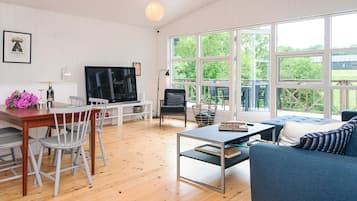 Uma sala de estar ampla com piso de madeira, uma mesa de jantar com cadeiras, um sofá, uma TV e grandes janelas com vista para o verde.
