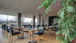 Uma sala de jantar moderna com várias mesas e cadeiras, janelas amplas e plantas em vasos.