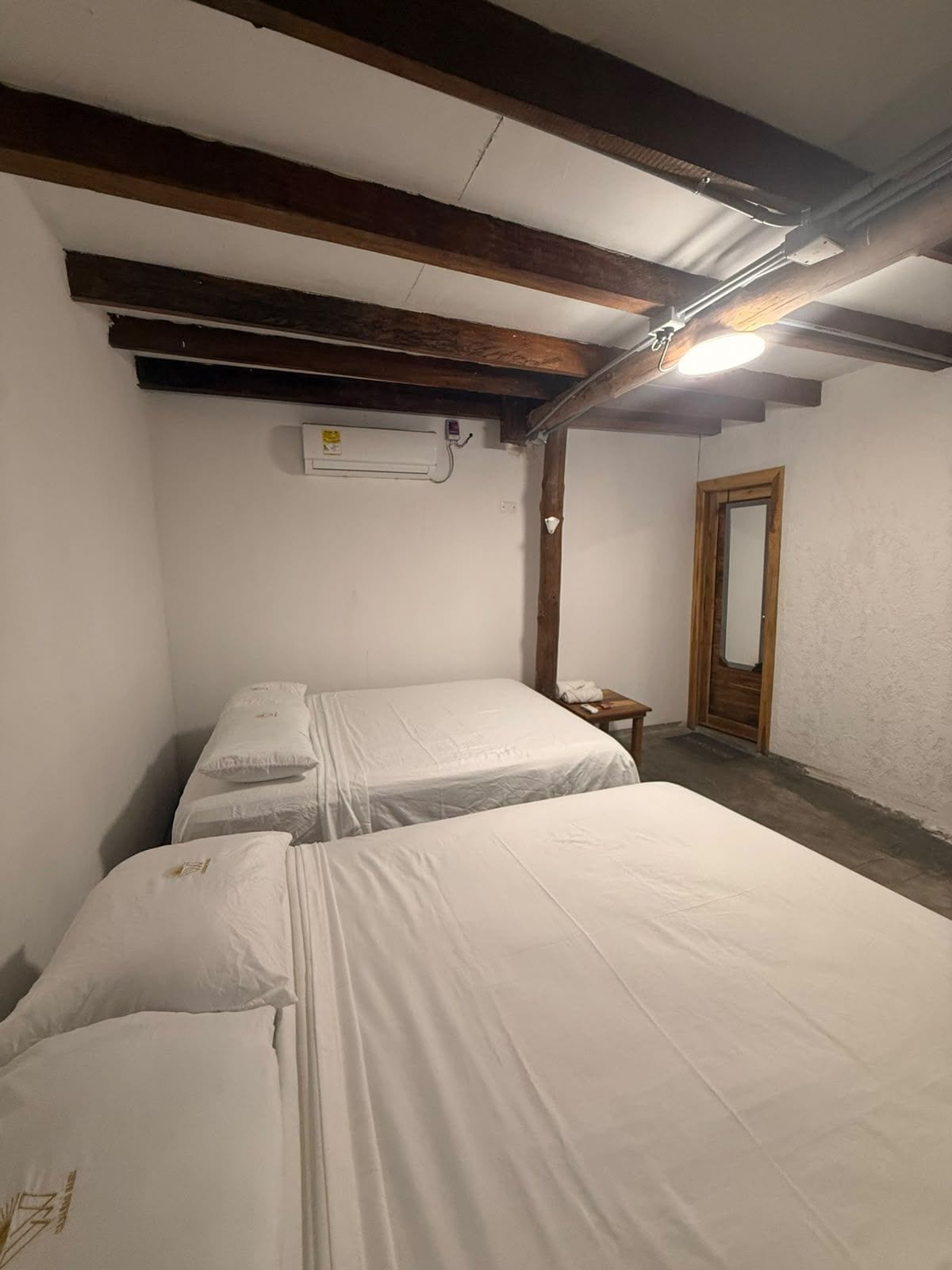 Quarto com duas camas, vigas de madeira no teto, uma mesa pequena e uma porta.