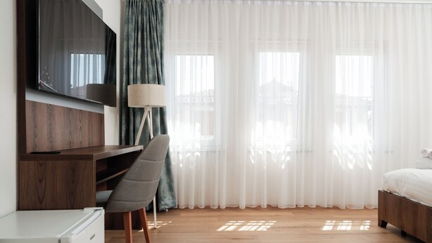 Quarto de hotel com cama, escrivaninha com cadeira, televisão e janela com cortinas finas.