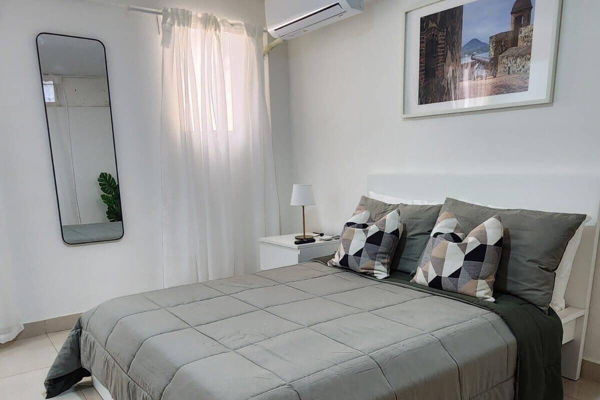 Quarto com cama, mesa de cabeceira com abajur, ar-condicionado fixado na parede, espelho e quadro emoldurado.
