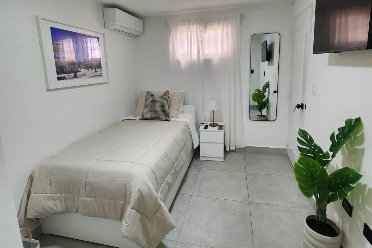 Quarto com cama, mesa de cabeceira, espelho, planta em vaso e um quadro na parede.