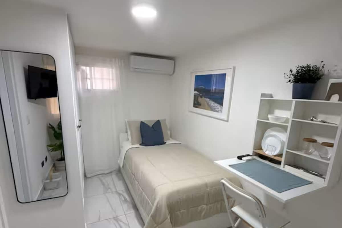 Quarto com cama, escrivaninha, cadeira, televisão e prateleira com objetos.