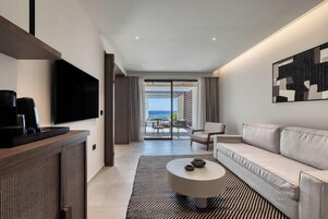 Quarto de hotel moderno com TV de tela plana, sofá, mesa de centro e vista para o oceano através de uma porta de vidro.