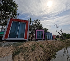 Três casas modulares com grandes janelas, localizadas à beira de um corpo d'água, com árvores e um céu claro ao fundo.