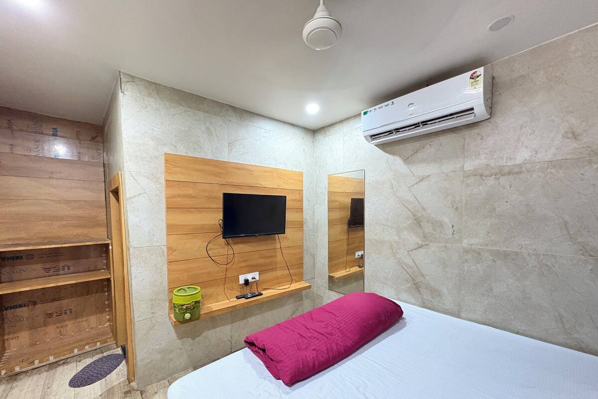 Quarto de hotel com parede de painel de madeira, televisão montada, cama com colchão branco e almofada rosa, lixeira verde pequena e unidade de ar condicionado.