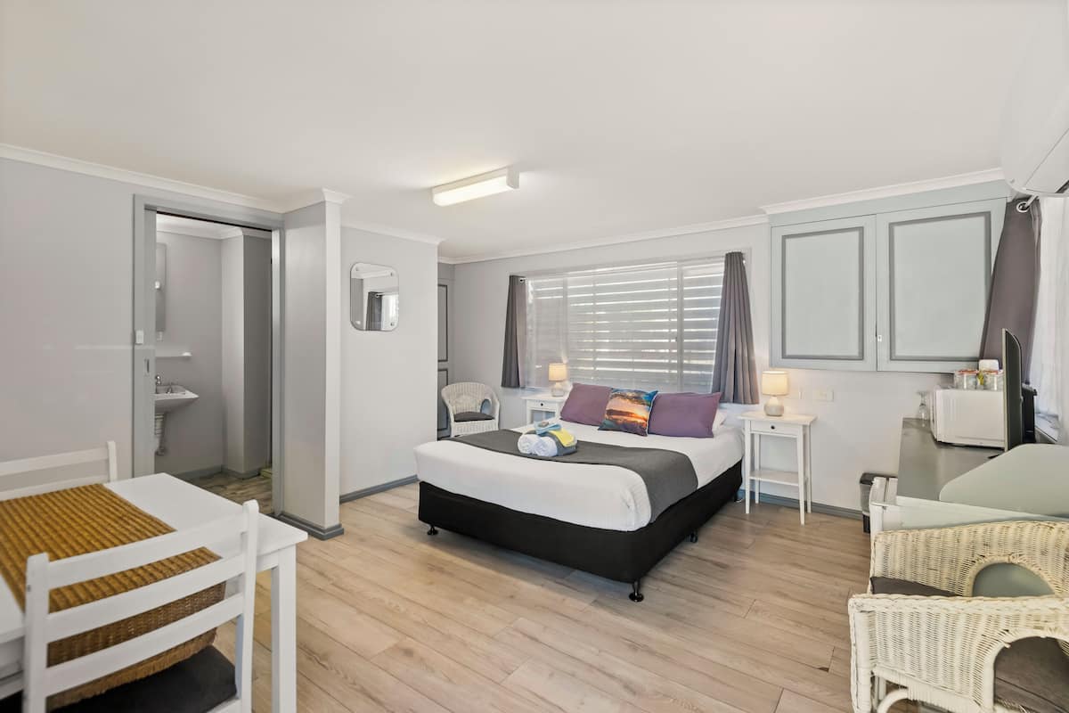 Quarto de hotel com cama, uma pequena mesa, uma cadeira, janela com persianas, espelho e guarda-roupa.