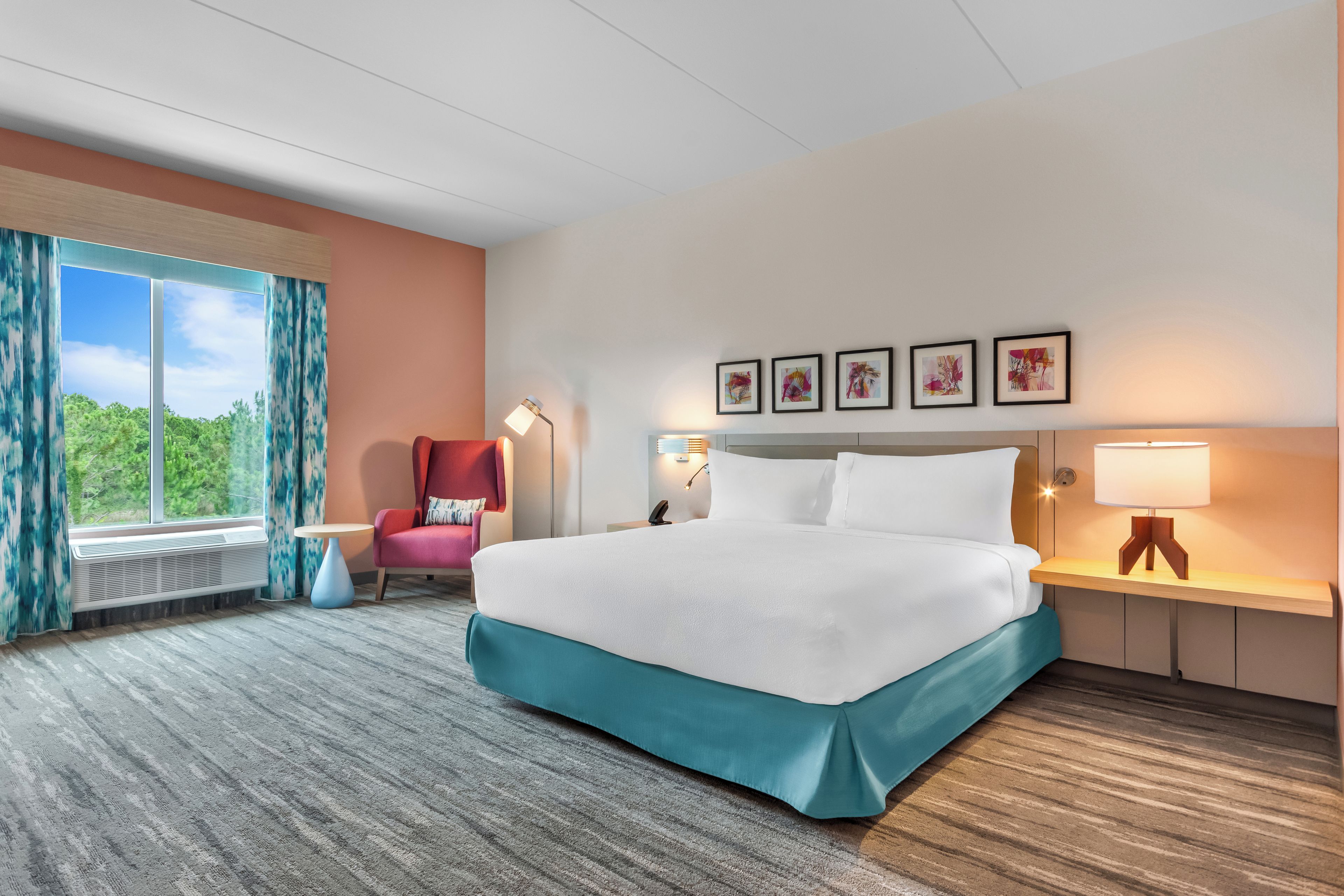 Quarto de hotel com cama grande, uma cadeira, escrivaninha e janela com cortinas.