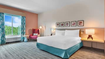 Quarto de hotel com cama grande, uma cadeira, escrivaninha e janela com cortinas.