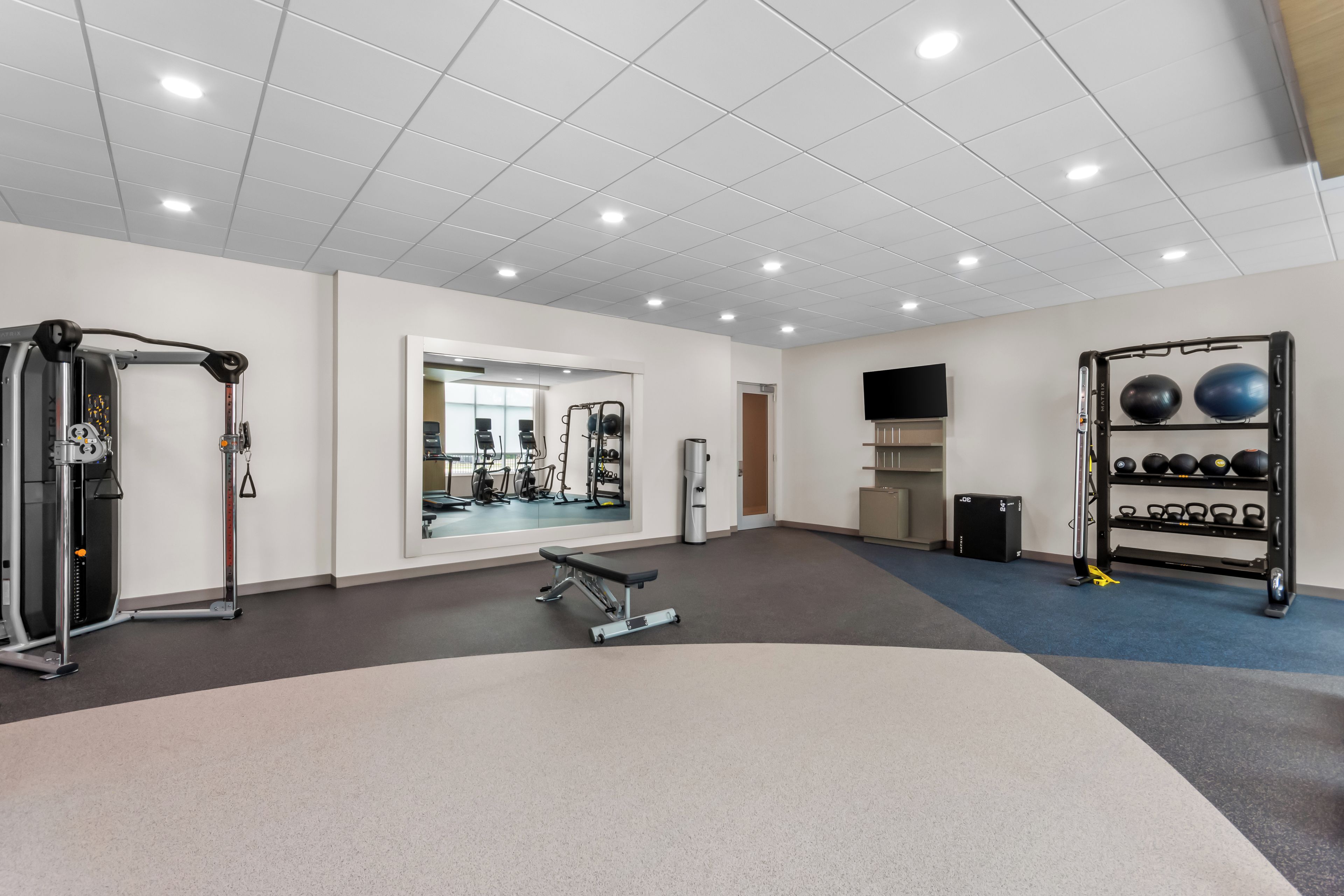 Academia com diversos equipamentos de exercícios, um banco e um espelho grande na parede.