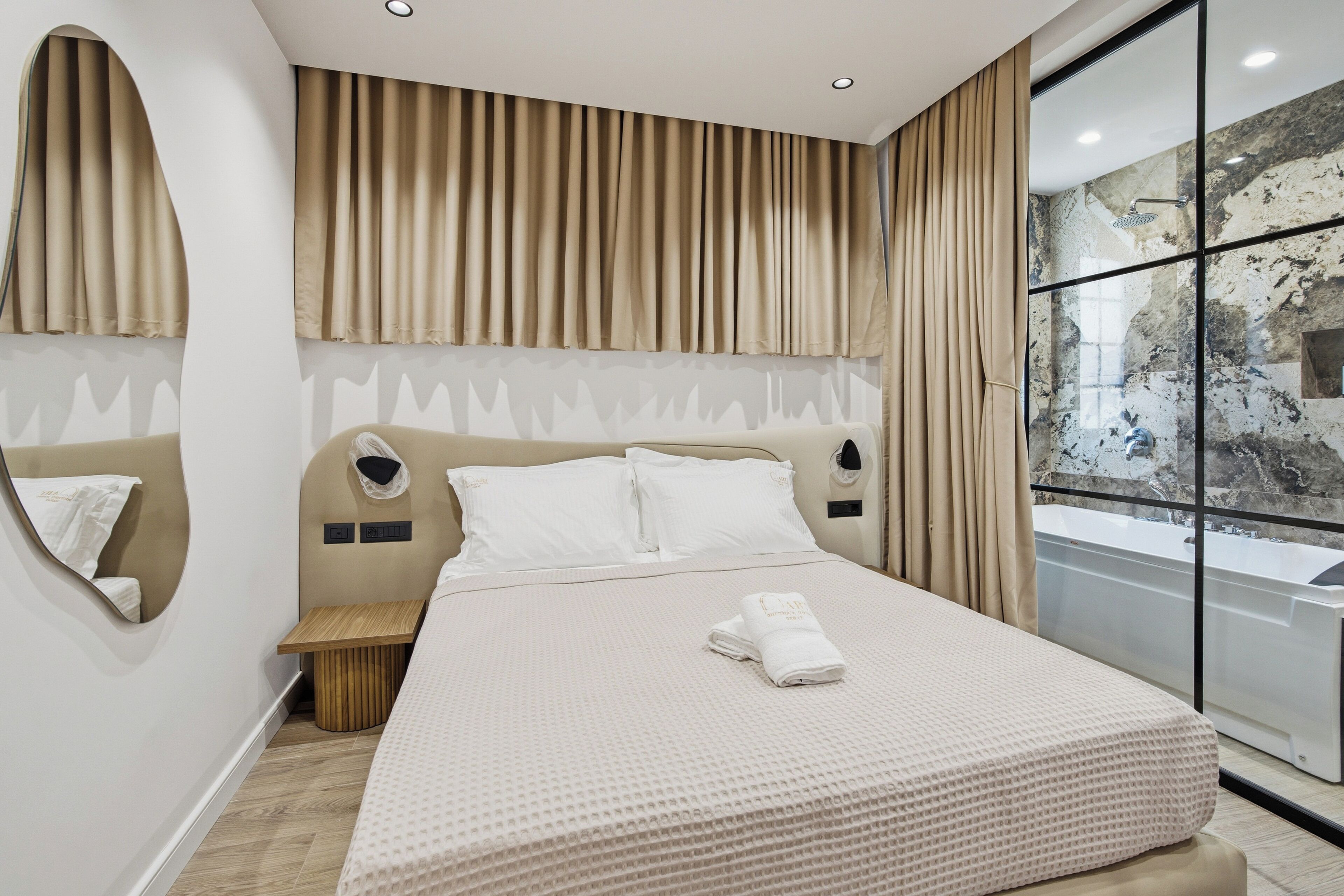 Quarto de hotel com uma cama grande, uma mesa de cabeceira pequena, um espelho e vista para uma banheira.