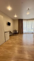Um quarto espaçoso com piso de madeira, televisão de tela plana na parede, mesa de jantar com cadeiras e uma janela grande com cortinas.