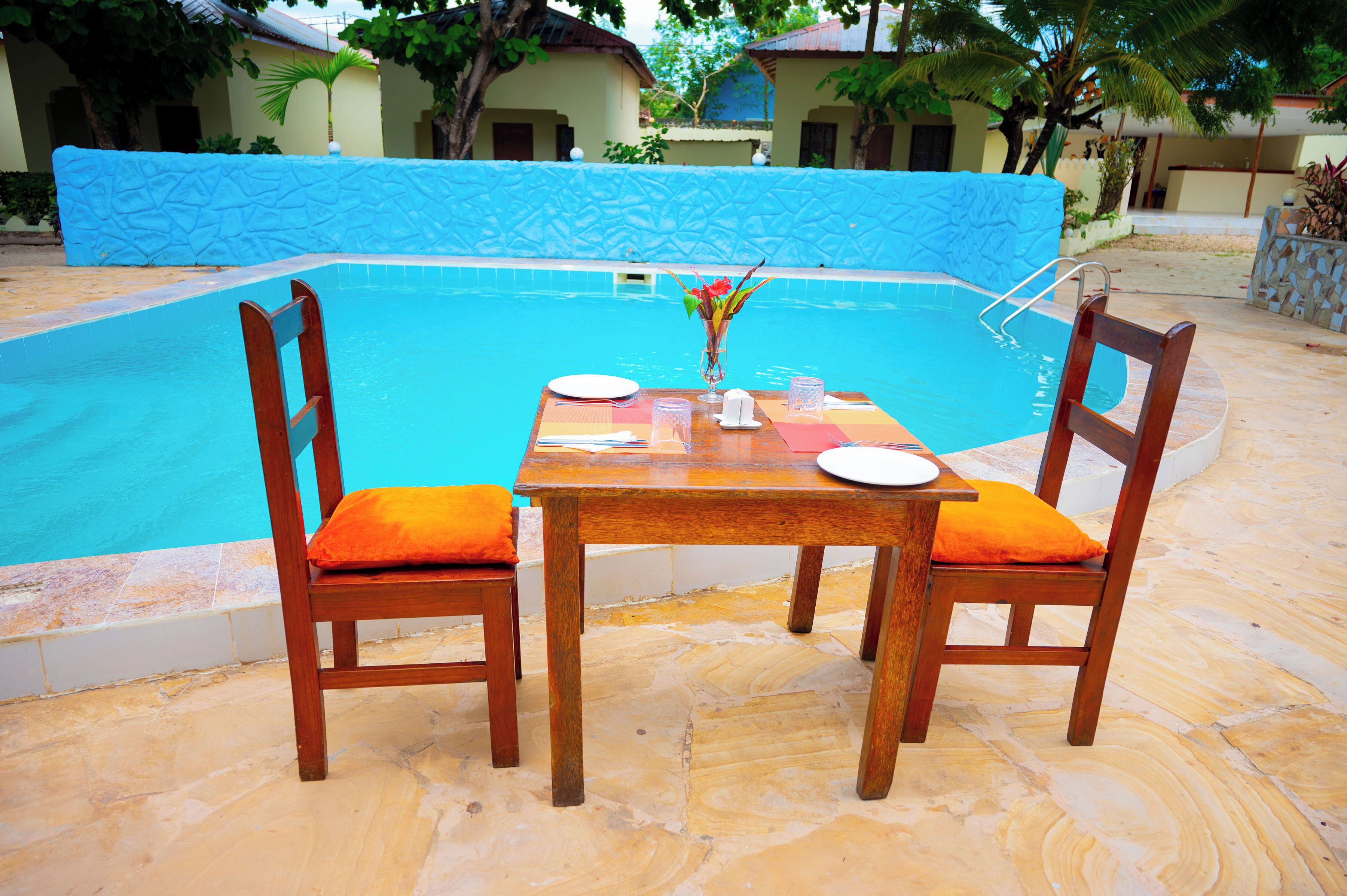 Uma área de refeições à beira da piscina com mesa e cadeiras, um vaso com flores e uma piscina azul cristalina.
