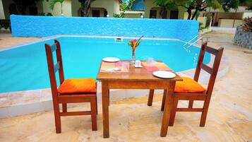 Uma área de refeições à beira da piscina com mesa e cadeiras, um vaso com flores e uma piscina azul cristalina.