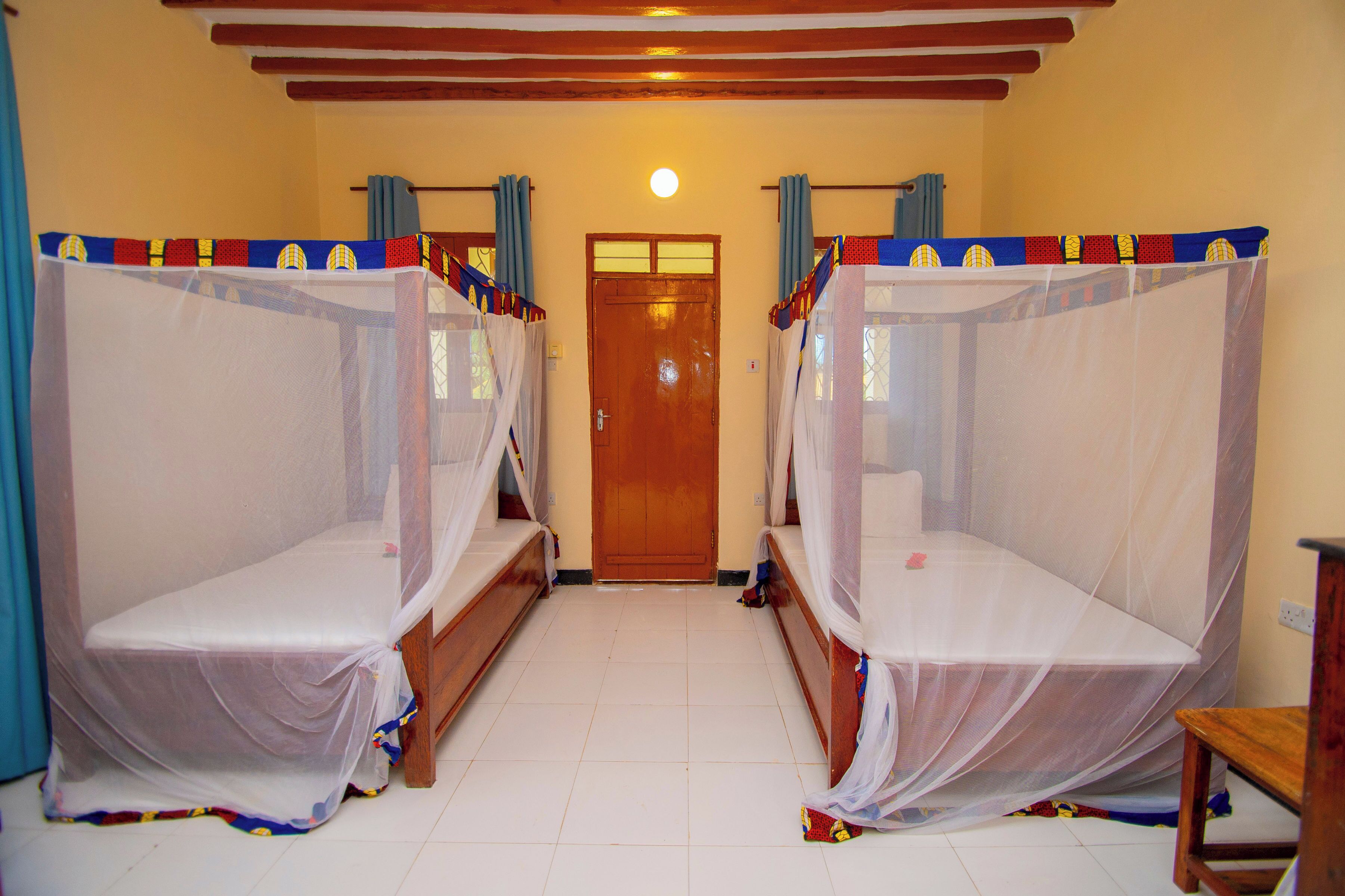 Quarto com duas camas de madeira, cada uma com mosquiteiro, uma porta de madeira e piso de azulejos.