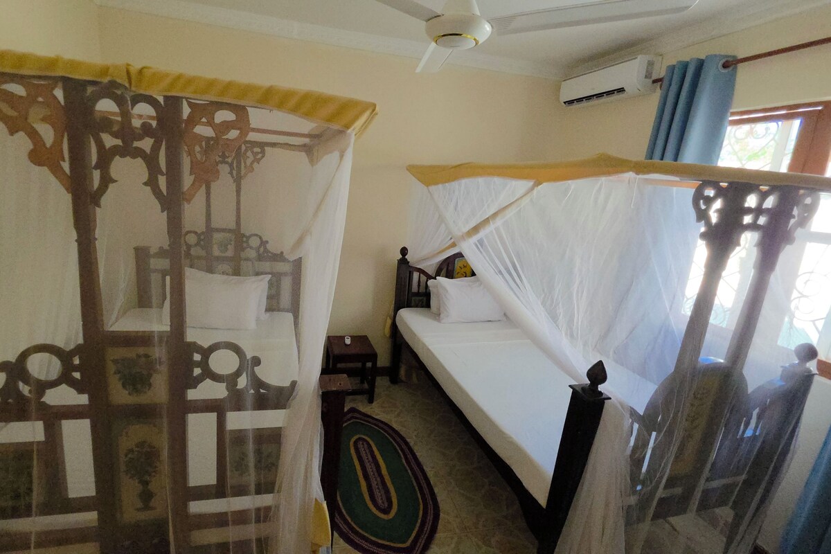 Quarto com duas camas, uma com dossel e outra sem, ventilador de teto, ar condicionado e janela com cortinas.