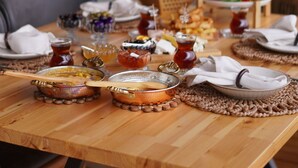 Mesa de jantar posta com diversos pratos e bebidas, uma cadeira e uma planta grande ao fundo.