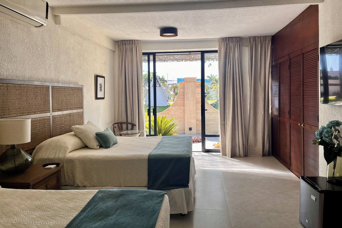 Quarto de hotel com duas camas, uma janela grande com cortinas, um ventilador de teto e vista para o exterior.