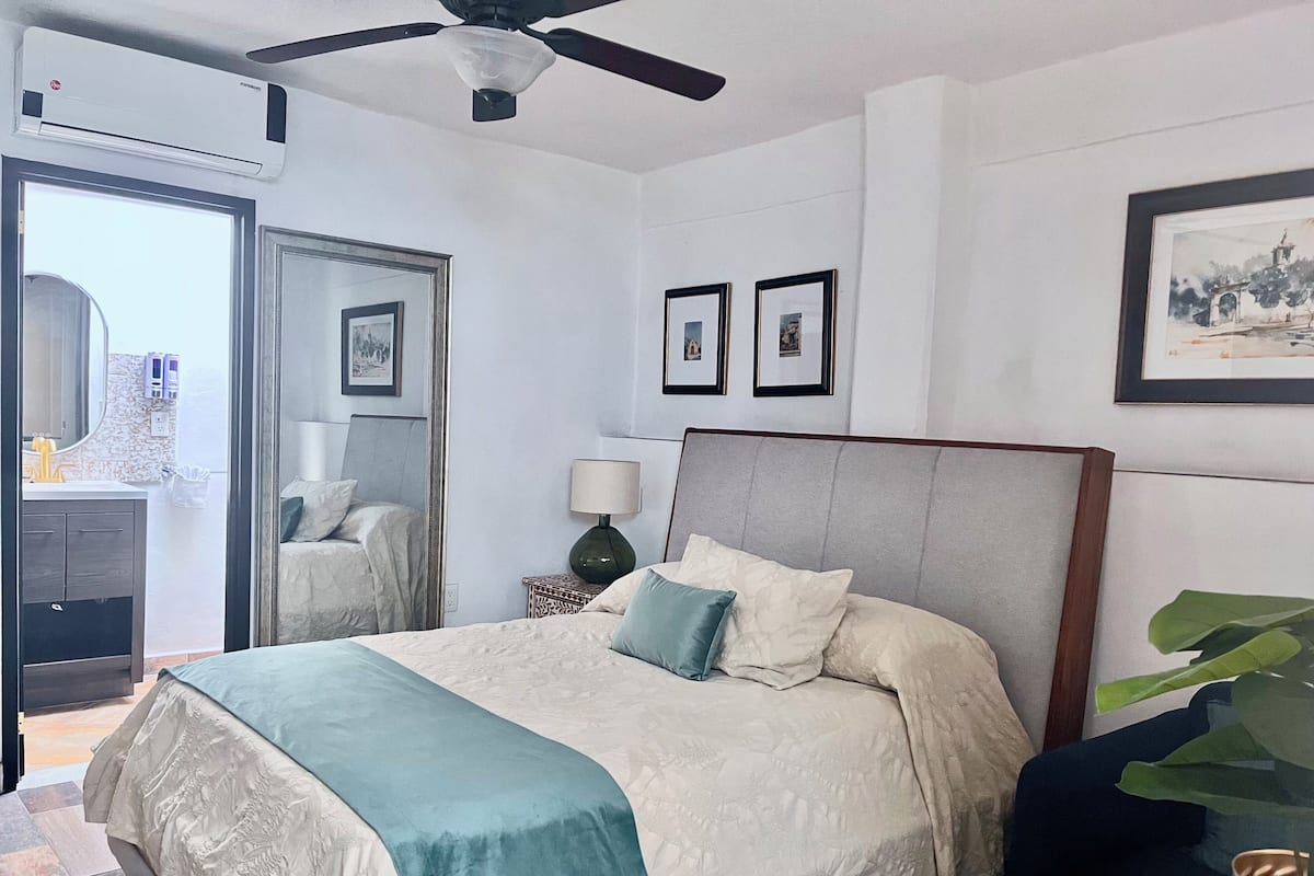 Quarto de hotel com cama, ventilador de teto, mesa de cabeceira com luminária, espelho e quadros nas paredes.