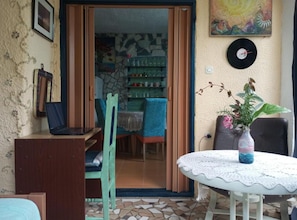 Um quarto com um laptop sobre uma escrivaninha de madeira, uma cadeira, uma mesa com toalha de renda, um vaso com flores e um disco de vinil na parede.